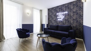 Uma sala de estar moderna com um sofá azul escuro, duas poltronas combinando e uma mesa de centro de vidro. É visível um grande mural com padrão de folha tropical e um letreiro de neon.