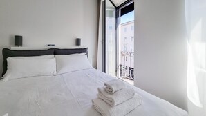 Quarto de hotel moderno com uma cama grande, roupa de cama branca, cabeceira escura e vista de um edifício por uma janela ampla.
