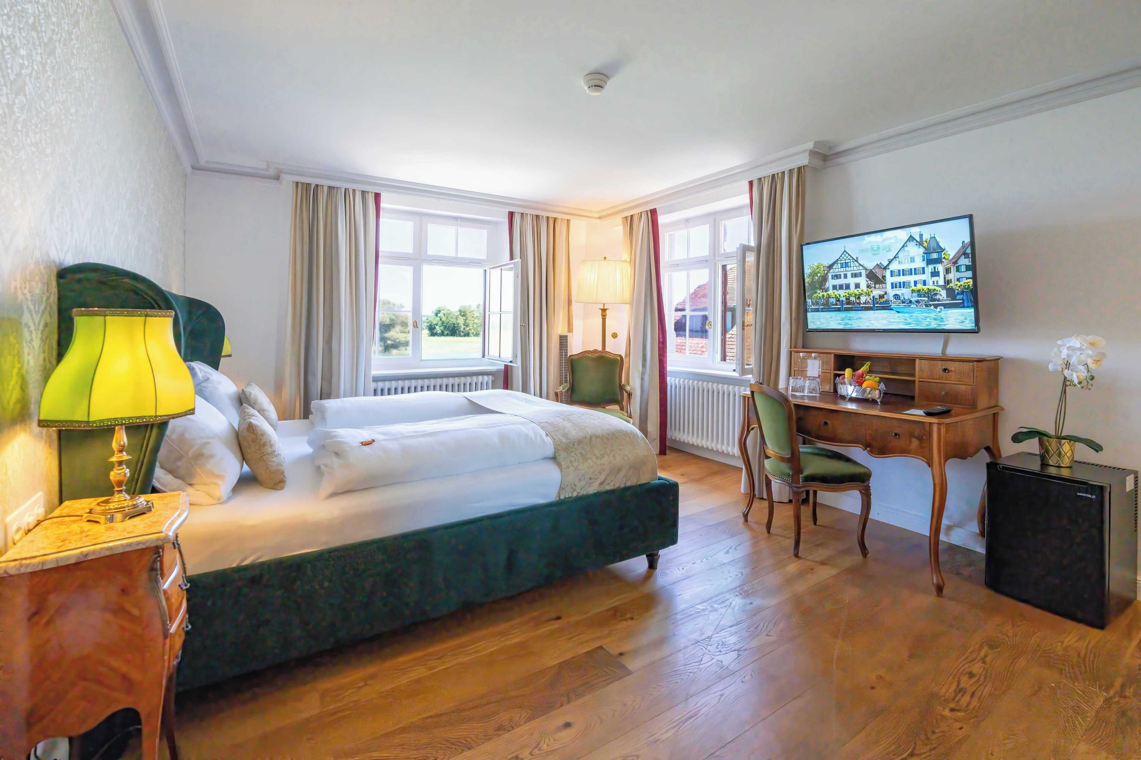 Quarto de hotel com cama, escrivaninha, cadeira, televisão e janela com cortinas.