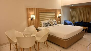 Quarto de hotel com uma cama grande, duas mesas de cabeceira, uma escrivaninha com cadeiras e uma poltrona azul.