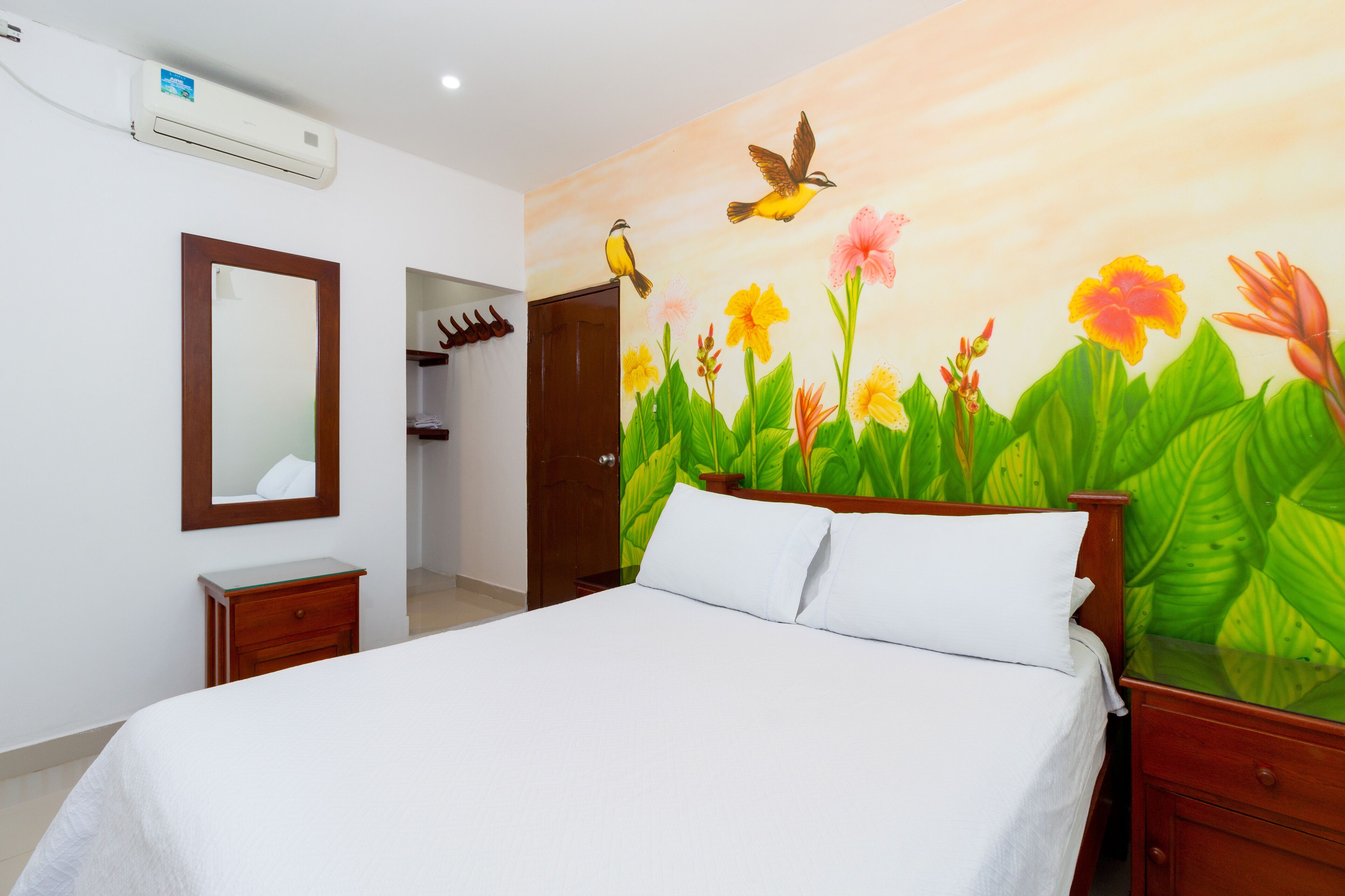 Quarto de hotel com cama, mesa de cabeceira de madeira, espelho e um mural na parede com pássaros e flores.