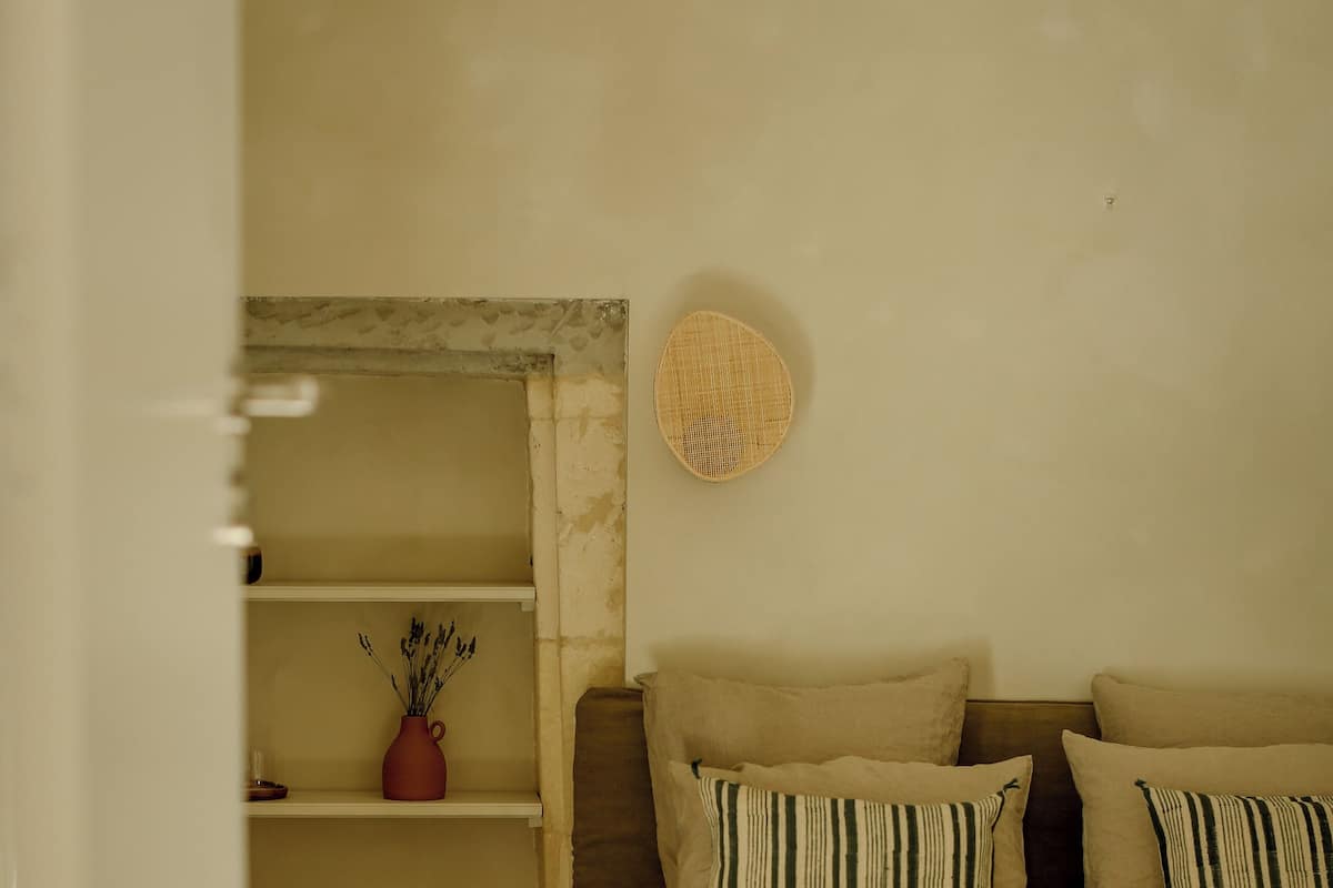 Quarto com cama, travesseiros, uma estante com um vaso e um objeto decorativo na parede.