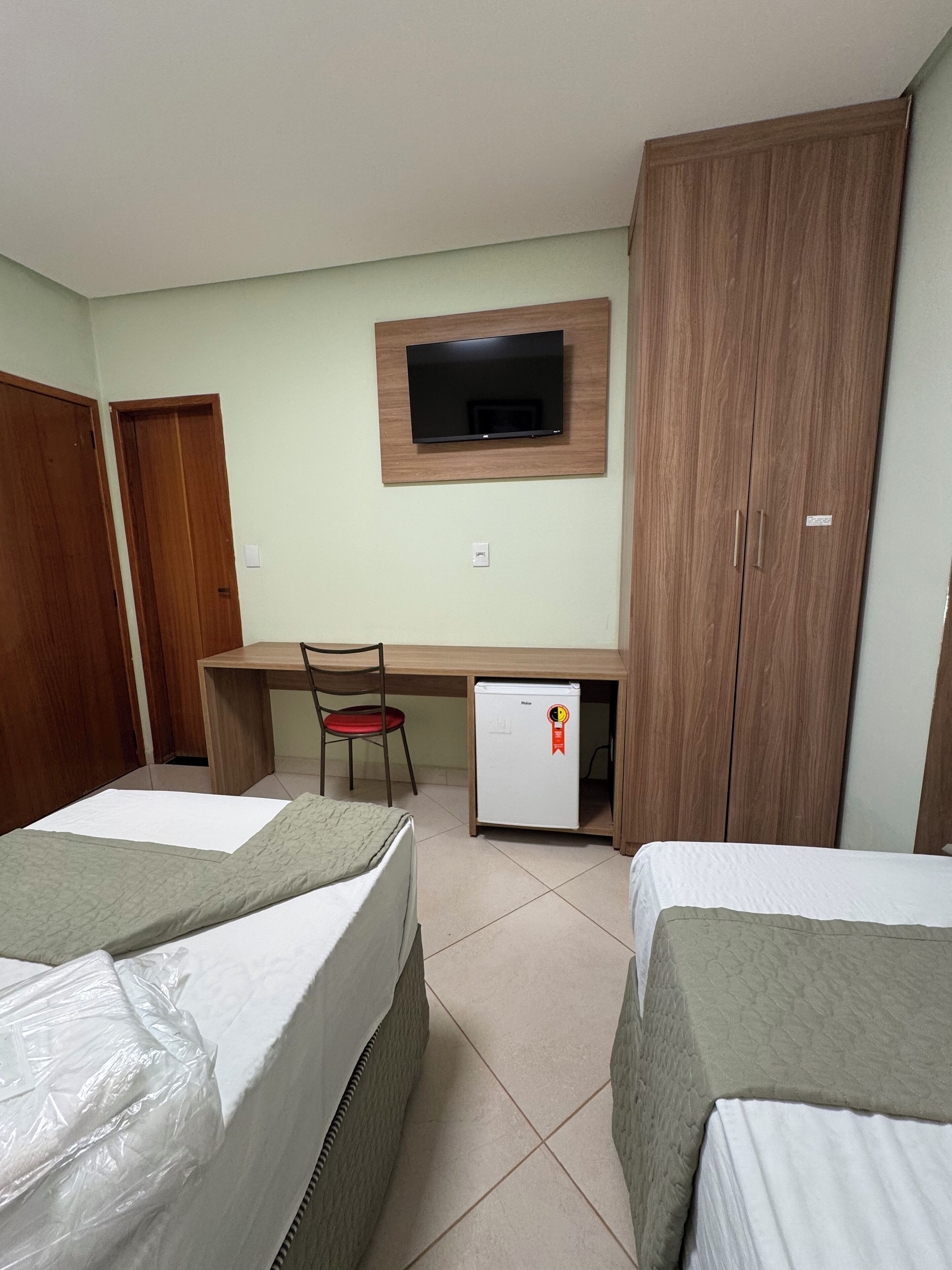 Quarto com duas camas, uma escrivaninha com cadeira, uma televisão fixada na parede e um frigorífico.