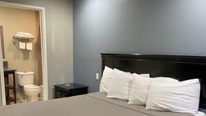 Quarto de hotel com uma cama grande, uma mesa de cabeceira, um banheiro visível através de uma porta aberta e uma luminária no teto.