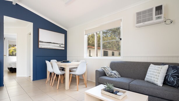 Uma sala de estar moderna com um sofá cinzento, uma mesa de jantar branca e cadeiras. Há uma janela com vista para árvores e um ar condicionado branco na parede.