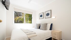 Quarto com uma cama grande, uma mesa de cabeceira, janela com vista para palmeiras e dois quadros pendurados na parede.