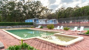 Piscina com duas áreas distintas, ladeada por um pátio de tijolos e cadeiras de lounge brancas.