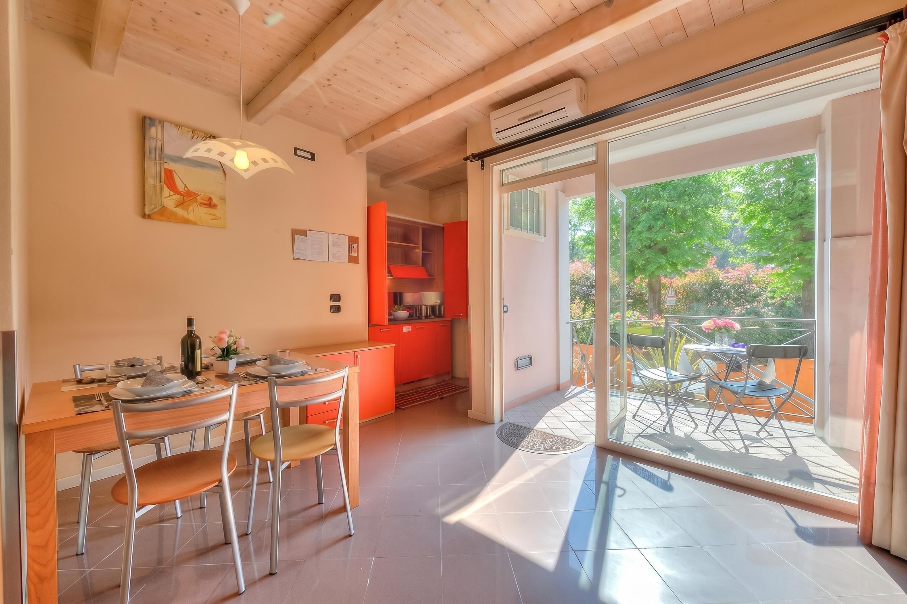 Uma área de refeições com uma mesa posta para quatro pessoas, uma cozinha com armários vermelhos e uma porta de vidro deslizante que dá para um pátio externo.