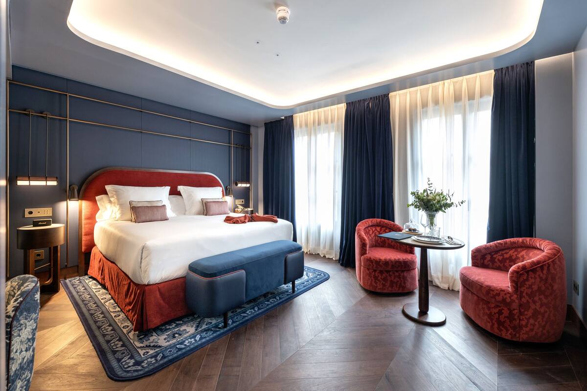 Quarto de hotel moderno com uma cama grande, área de estar com duas poltronas vermelhas, uma mesinha redonda e uma janela ampla com cortinas.