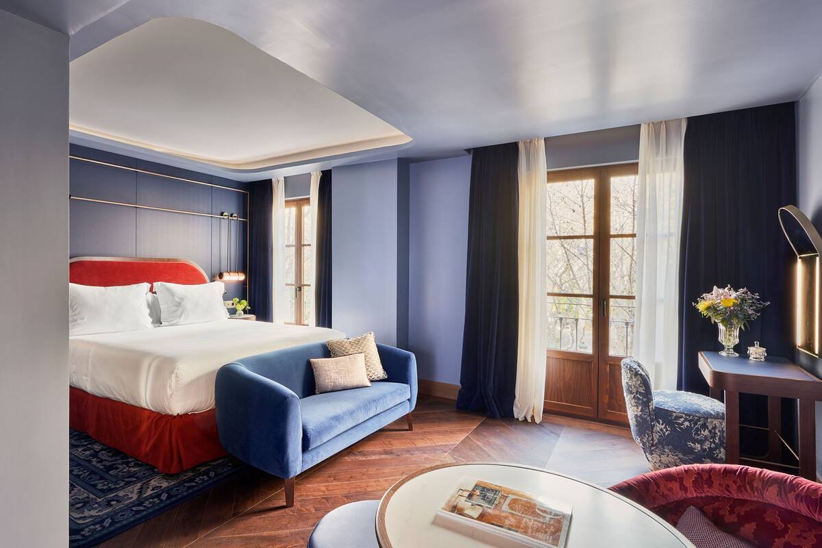 Quarto de hotel moderno com cama, sofá azul, mesa redonda e escrivaninha com abajur.