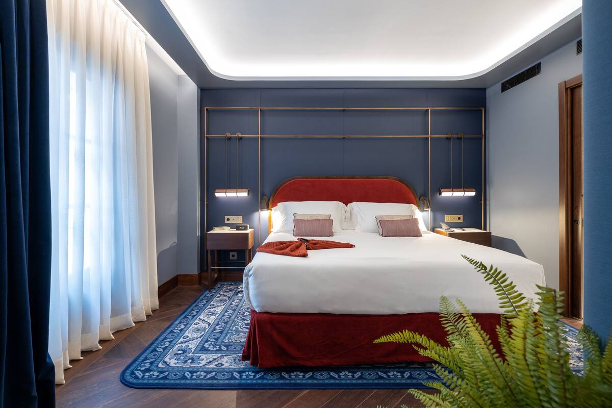 Quarto de hotel moderno com uma cama grande, duas mesinhas de cabeceira, um criado-mudo, um tapete azul estampado e uma janela ampla com cortinas leves.