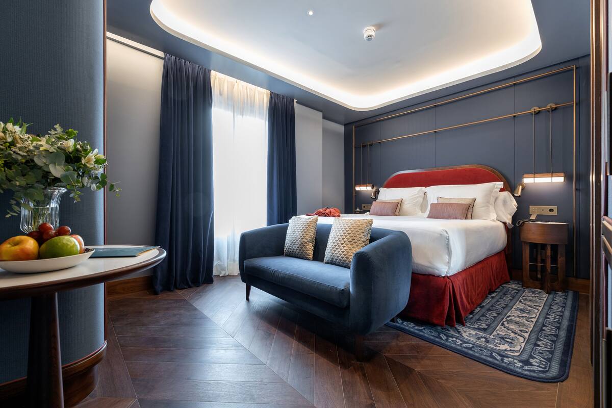 Quarto de hotel moderno com uma cama grande, um sofá azul escuro, uma mesinha de centro com frutas e flores, e uma janela ampla com cortinas.