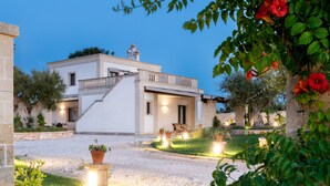 Uma casa moderna com fachada branca, varanda e um jardim bem iluminado com vasos e mobiliário de exterior.
