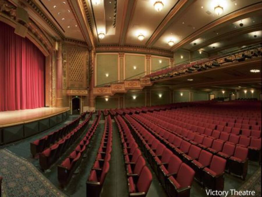 Um teatro com assentos vermelhos, palco e detalhes arquitetônicos elaborados.
