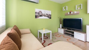 Sala de estar com parede verde, televisão de tela plana, sofá branco e pequena mesa branca com uma vela.