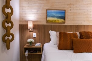Quarto de hotel com cabeceira de madeira, pintura de uma praia, mesa de cabeceira com telefone e um vaso de flores, e um grande painel de parede trançado marrom.