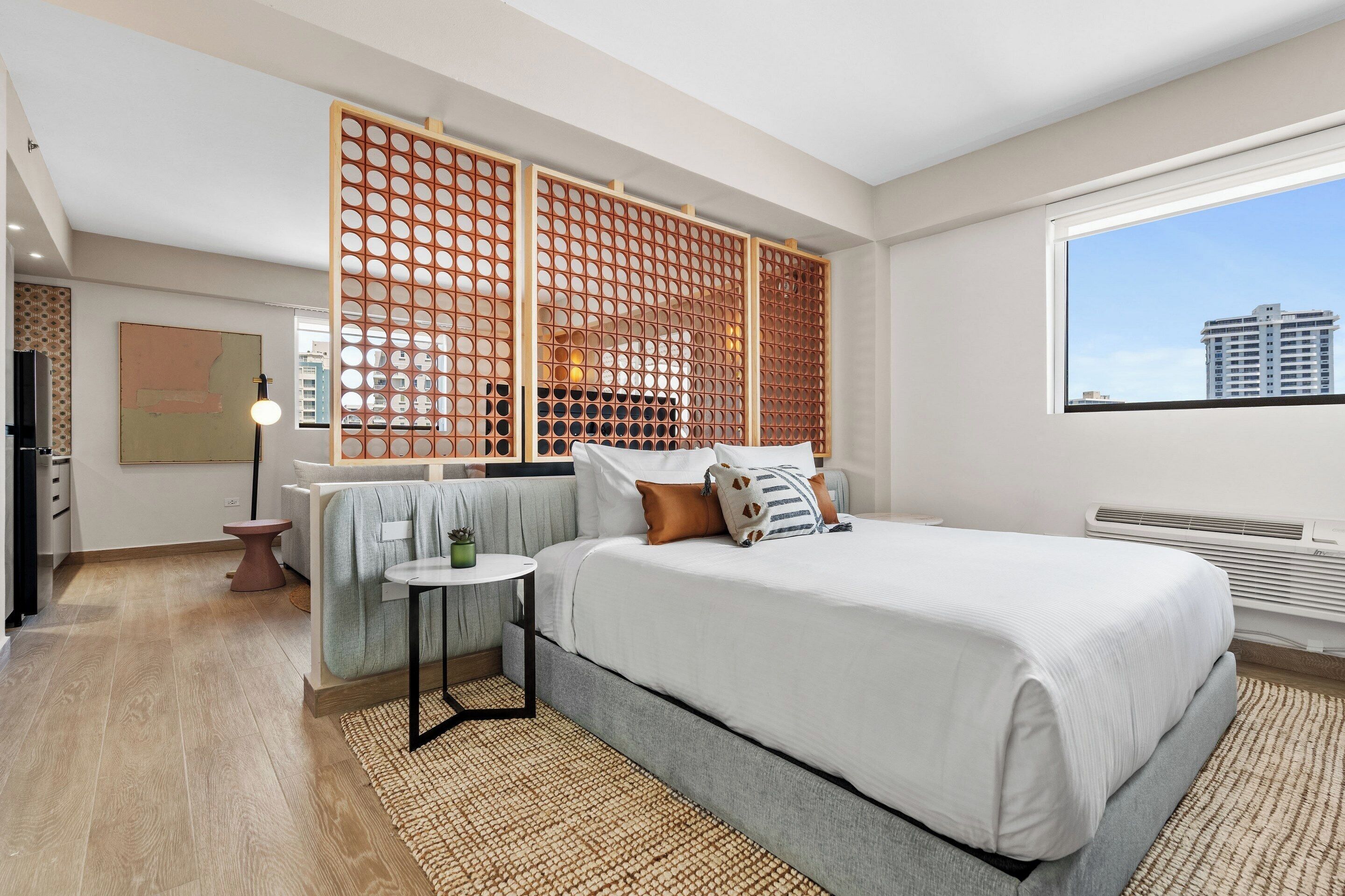 Quarto moderno com uma cama grande, uma mesinha, um biombô decorativo e vista para a cidade através de uma janela.