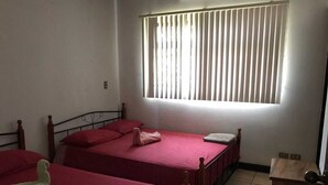 Quarto com duas camas de solteiro, janela com persianas, pequena mesa de madeira e piso ladrilhado.