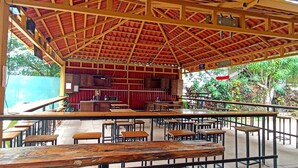 Uma área de refeições ao ar livre com mesas e bancos de madeira, um palco com alto-falantes e uma parede vermelha ao fundo.