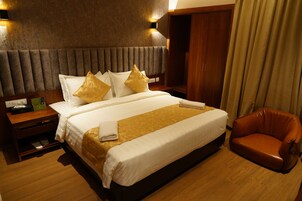 Quarto de hotel com uma cama grande, cabeceira de madeira, poltrona de couro marrom e mesa de cabeceira de madeira com abajur.