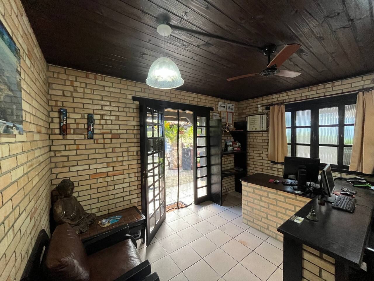 Quarto com parede de tijolos, ventilador de teto, mesa com um computador e porta que dá para o exterior.