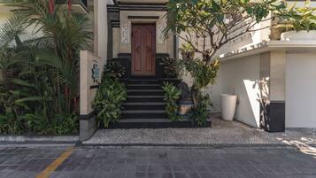 Entrada de um edifício com porta de madeira, degraus de acesso e um pequeno jardim com plantas.