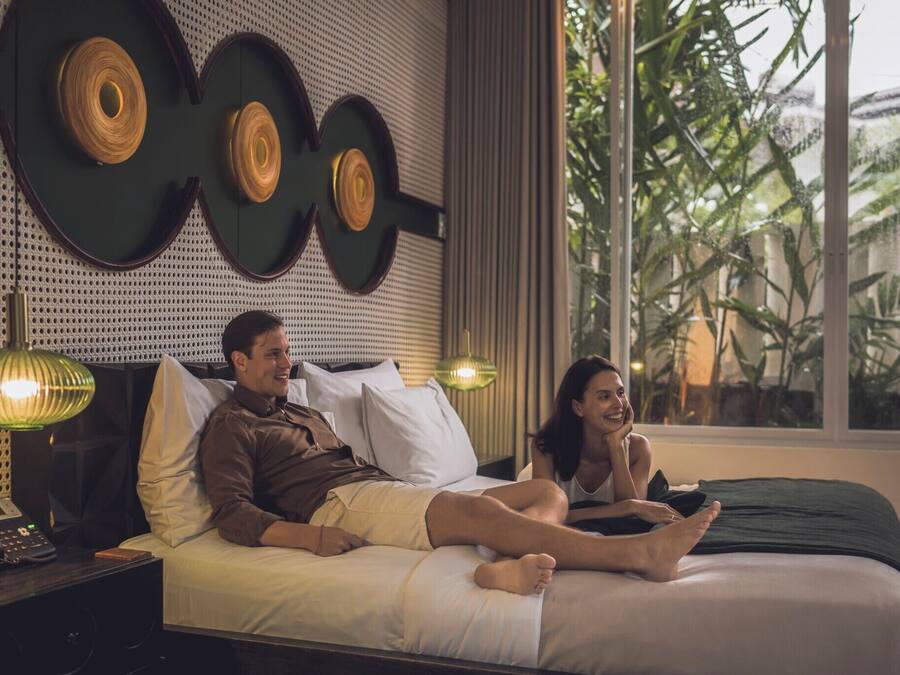 Quarto com cama, duas luminárias de cabeceira, mesa de cabeceira com telefone e janela com vista para área verde.