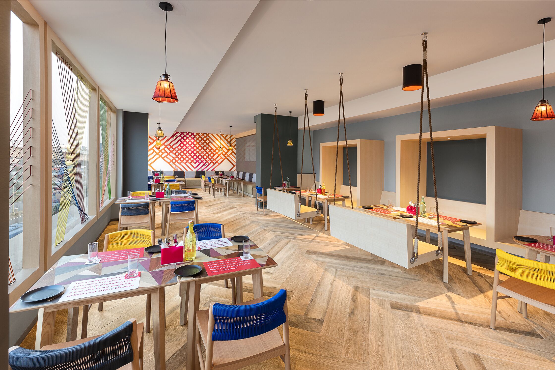Restaurante moderno com chão de madeira, cadeiras coloridas e luminárias pendentes. Mesas montadas com cardápios e copos.