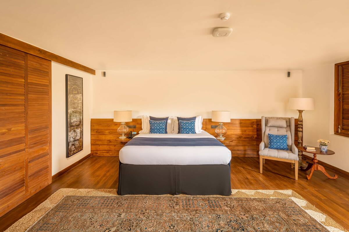 Quarto com uma cama grande, cabeceira de madeira, duas mesas de cabeceira com abajures, uma cadeira e um tapete.