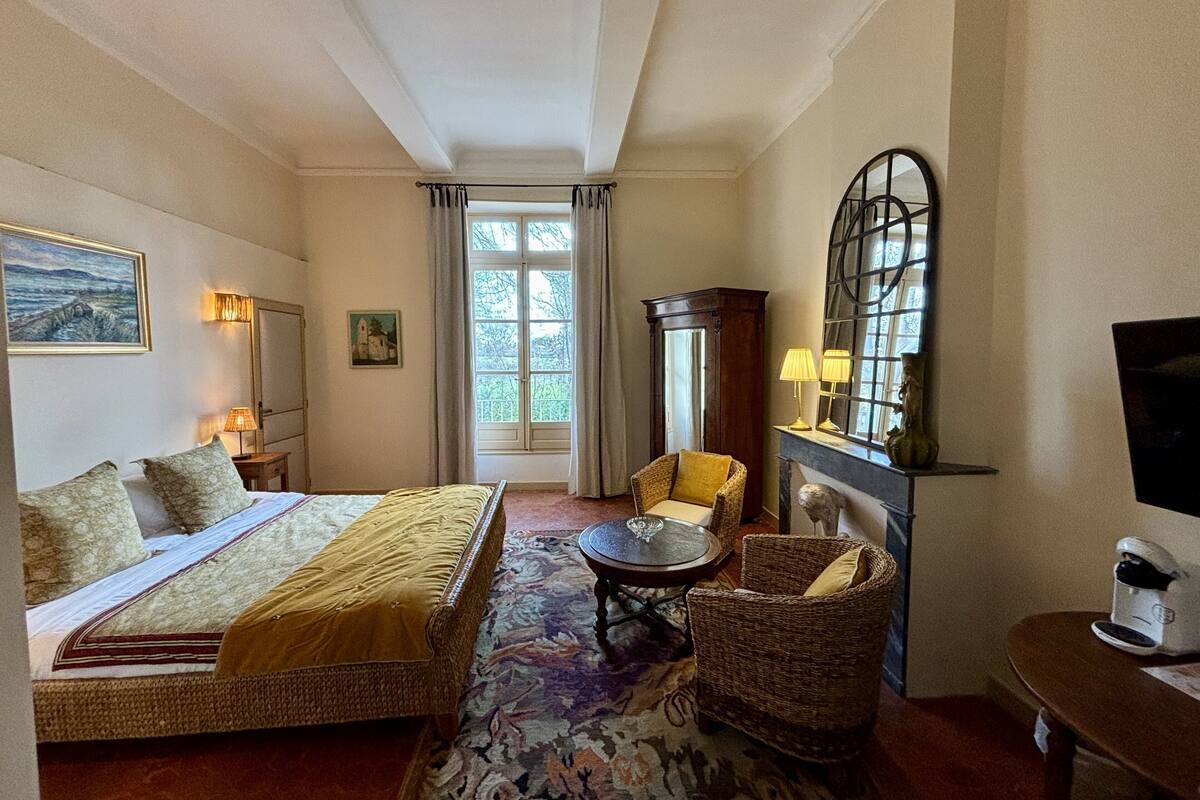 Quarto de hotel com cama, sofá, mesa e cadeira. Há uma janela com cortinas, um quadro na parede e um abajur.
