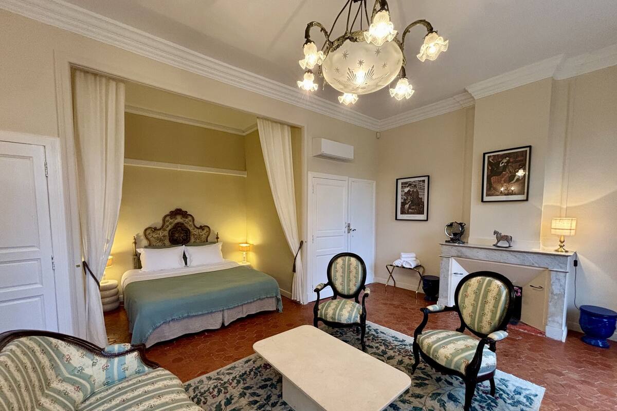 Um quarto com uma cama, duas poltronas, uma mesinha, lareira e um candelabro.