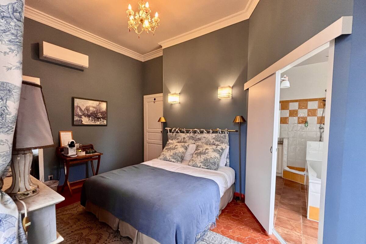 Quarto com cama, abajures, mesa de cabeceira, ar condicionado fixado na parede, pintura e porta para um banheiro.