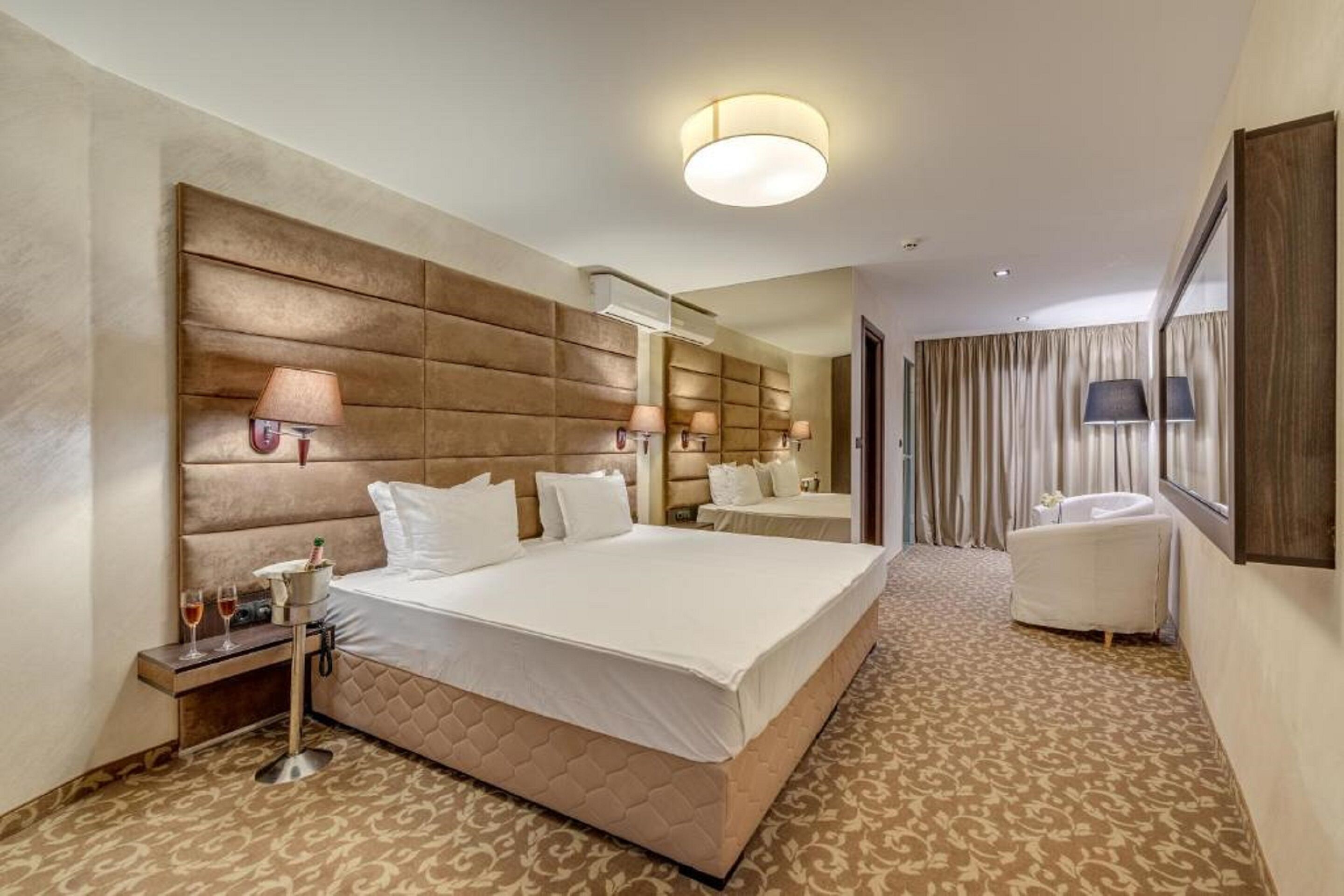 Quarto de hotel com uma cama grande, luminárias de cabeceira, uma mesinha com uma garrafa e copos, uma cadeira e um espelho grande.