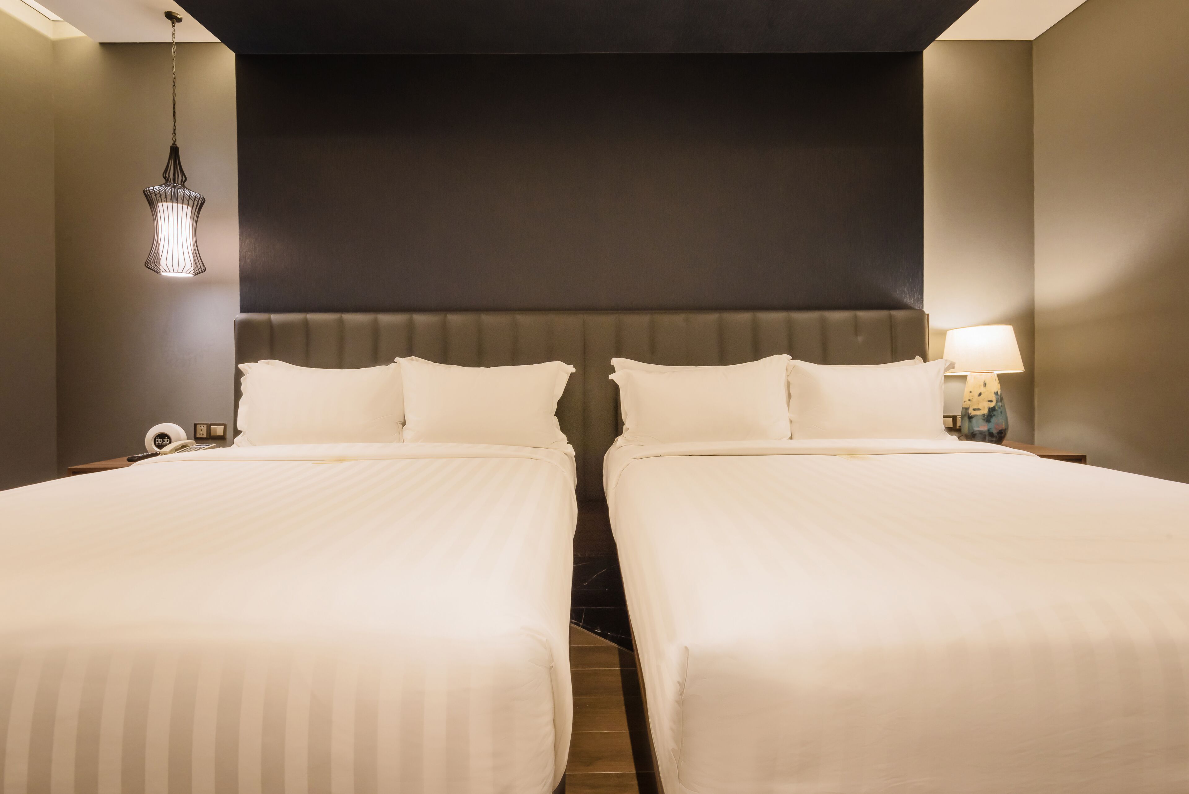 Quarto de hotel com duas camas, cabeceira escura, luminária pendente e mesa de cabeceira com abajur.