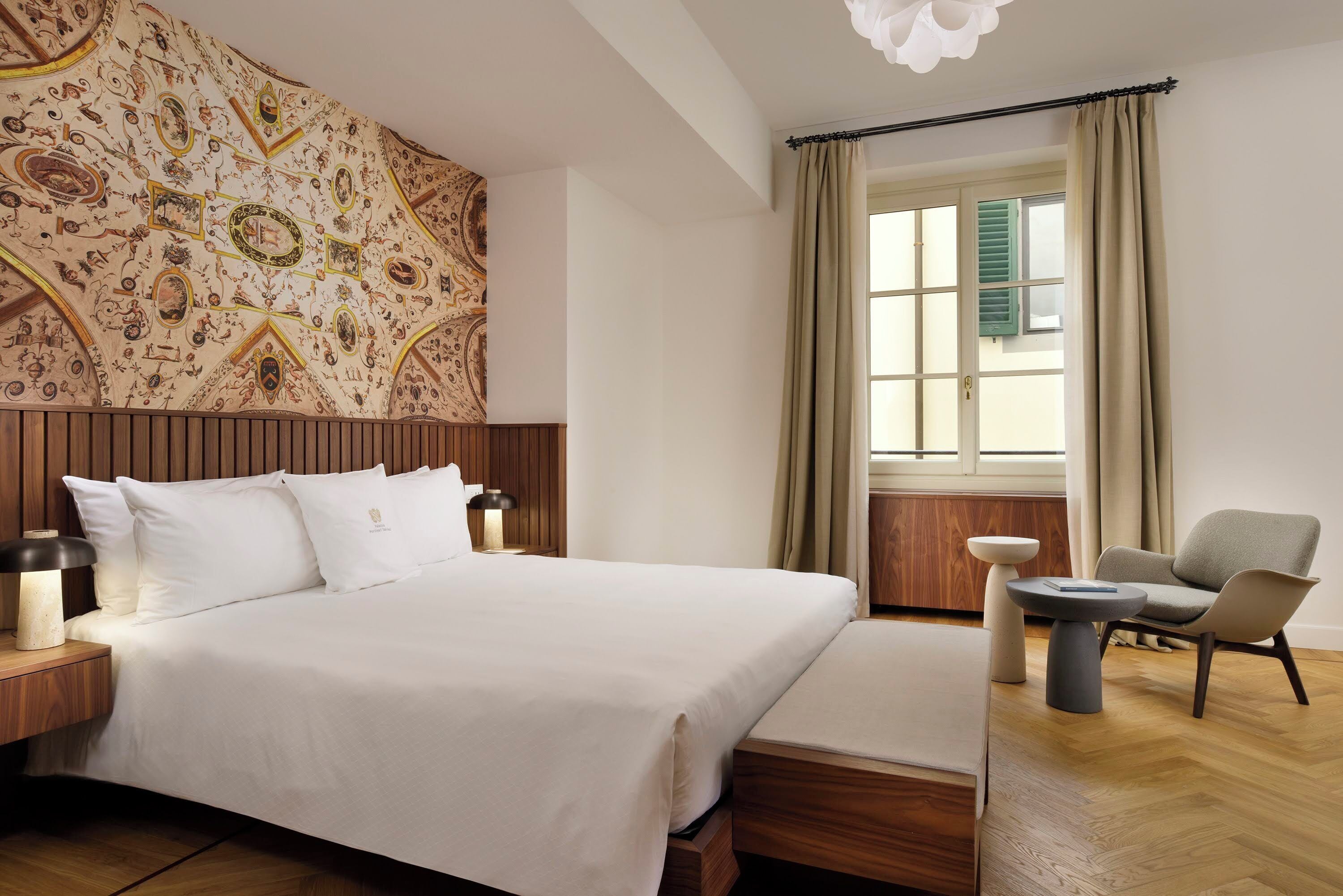 Quarto de hotel com cama grande, cabeceira de madeira, mesa de cabeceira com abajur, mesa redonda pequena, uma cadeira e uma janela com cortinas.