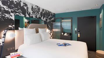 Quarto de hotel moderno com uma cama grande, mesa de cabeceira com abajur, espelho e porta com puxador.