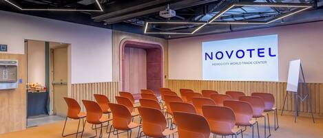 Sala de conferências com fileiras de cadeiras laranja, uma tela de projeção exibindo 'NOVOTEL' e uma lousa.