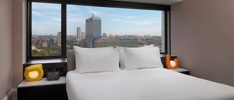 Quarto de hotel com uma grande janela que oferece vista para a cidade, uma cama com roupa de cama branca, uma mesa de cabeceira com abajur e uma pequena mesa com um dispositivo.