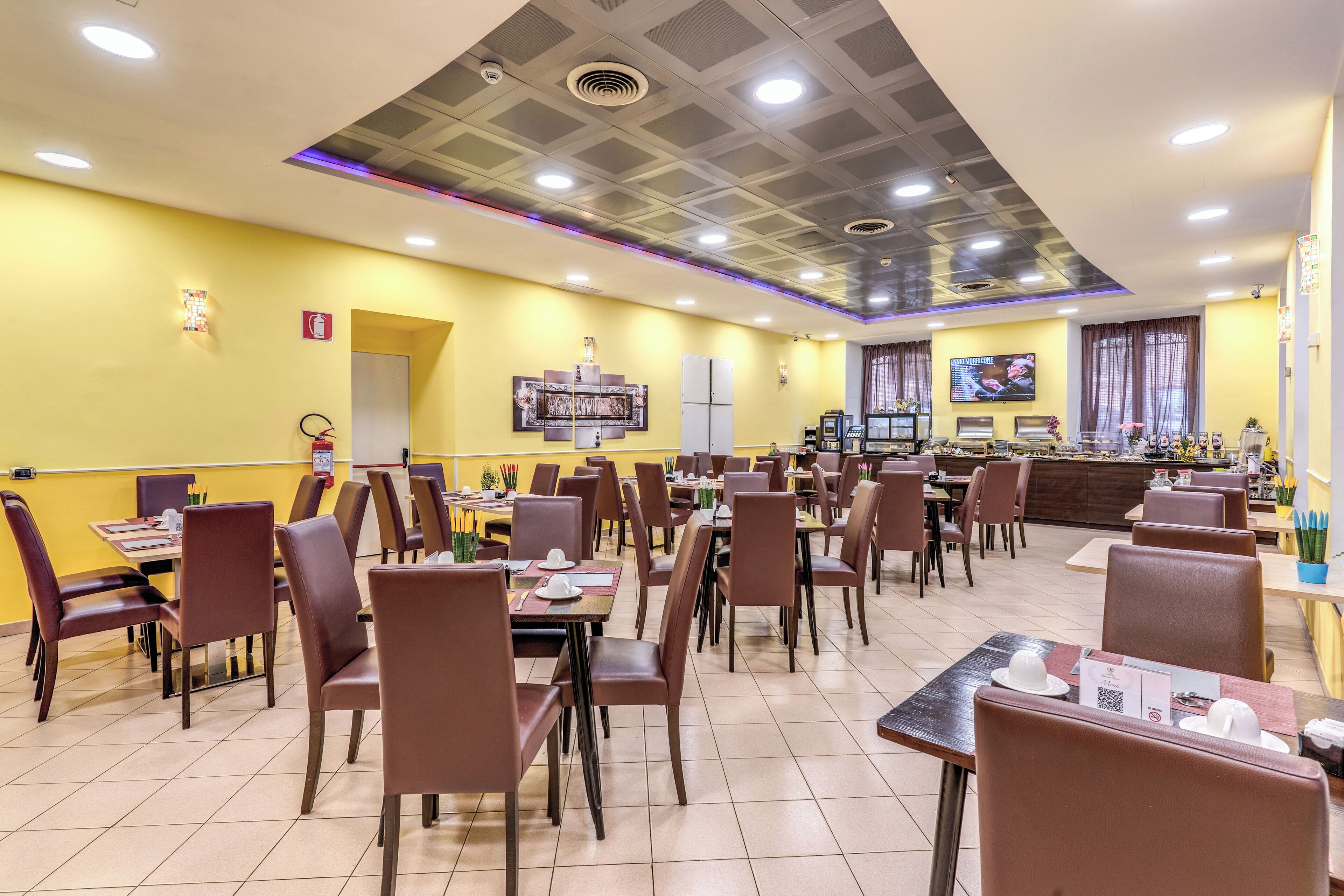 Um restaurante com várias mesas e cadeiras, um balcão com cardápio e uma tela de televisão na parede.