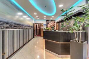 Uma área de recepção de hotel com design moderno, com um balcão de atendimento, uma grande planta em vaso e um mural com vista para a cidade.