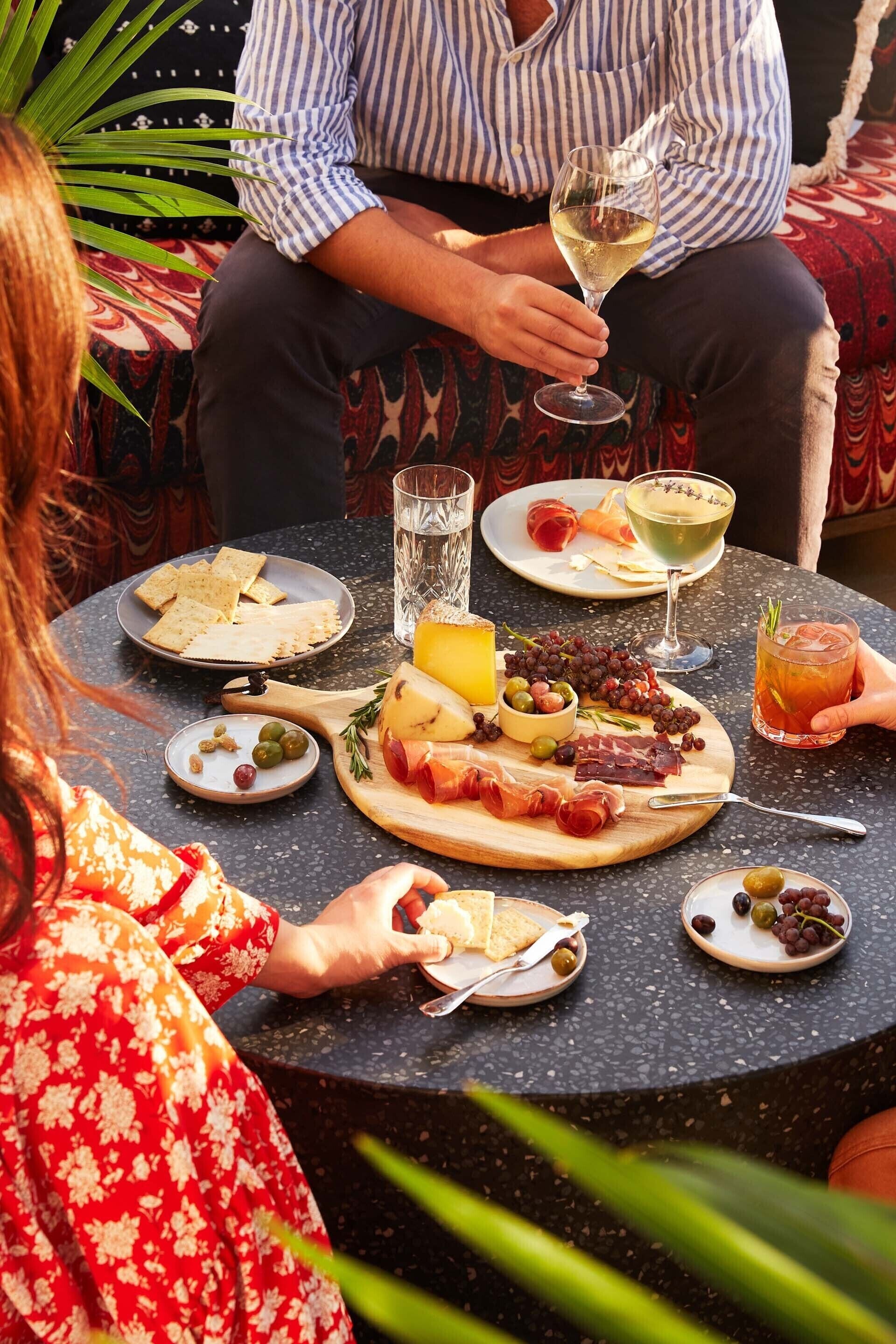 Uma mesa redonda com diversos alimentos e bebidas, incluindo queijo, carnes e azeitonas.