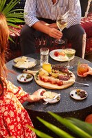 Uma mesa redonda com diversos alimentos e bebidas, incluindo queijo, carnes e azeitonas.
