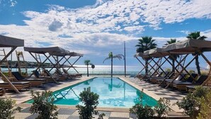Uma área de piscina com espreguiçadeiras e guarda-sóis, céu claro e vista para o mar.