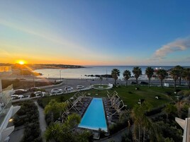 Piscina com espreguiçadeiras, vista para o oceano e um pôr do sol.