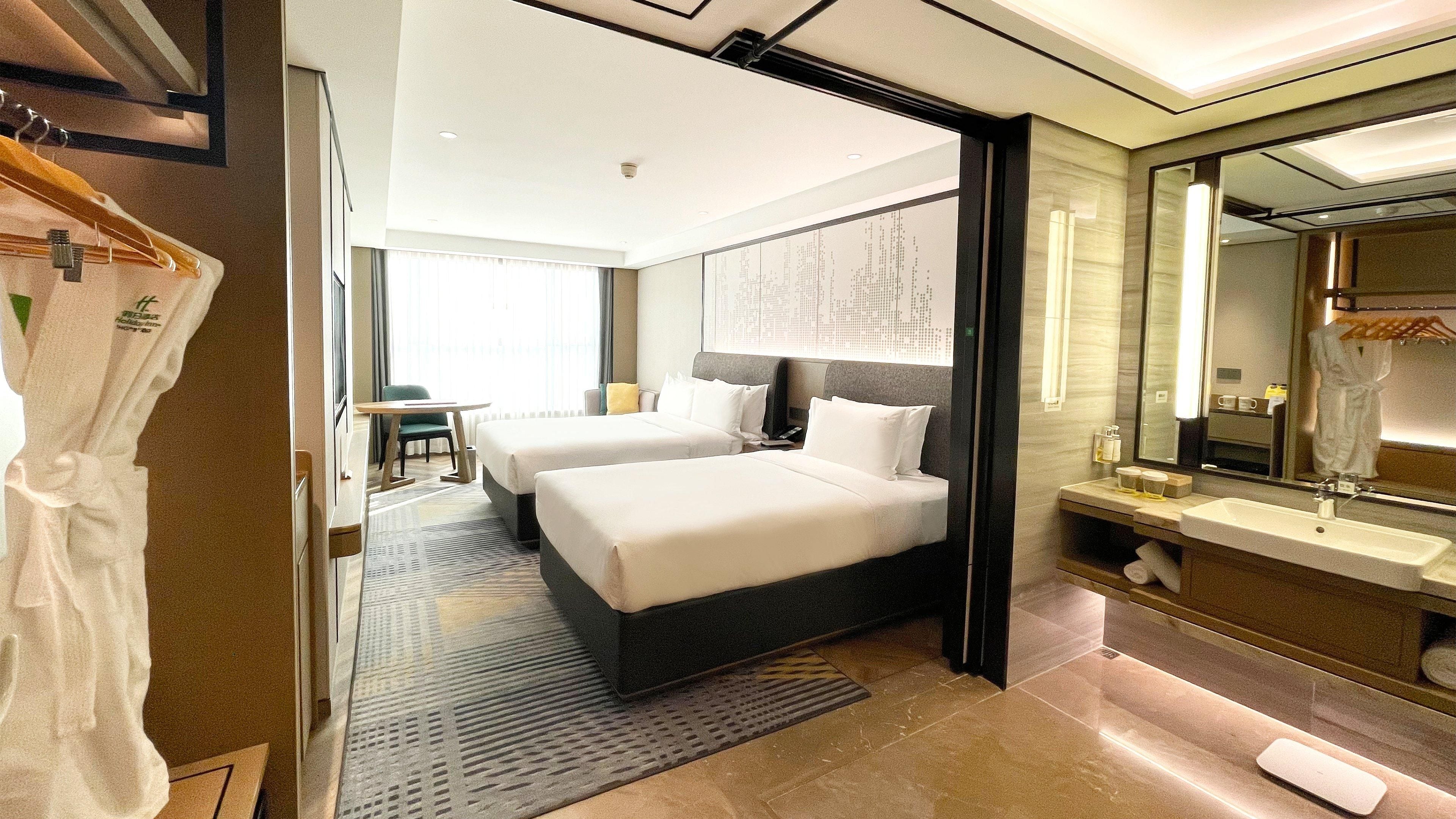 Quarto de hotel com duas camas, uma escrivaninha, uma cadeira, um espelho grande e banheiro com pia e porta-toalhas.
