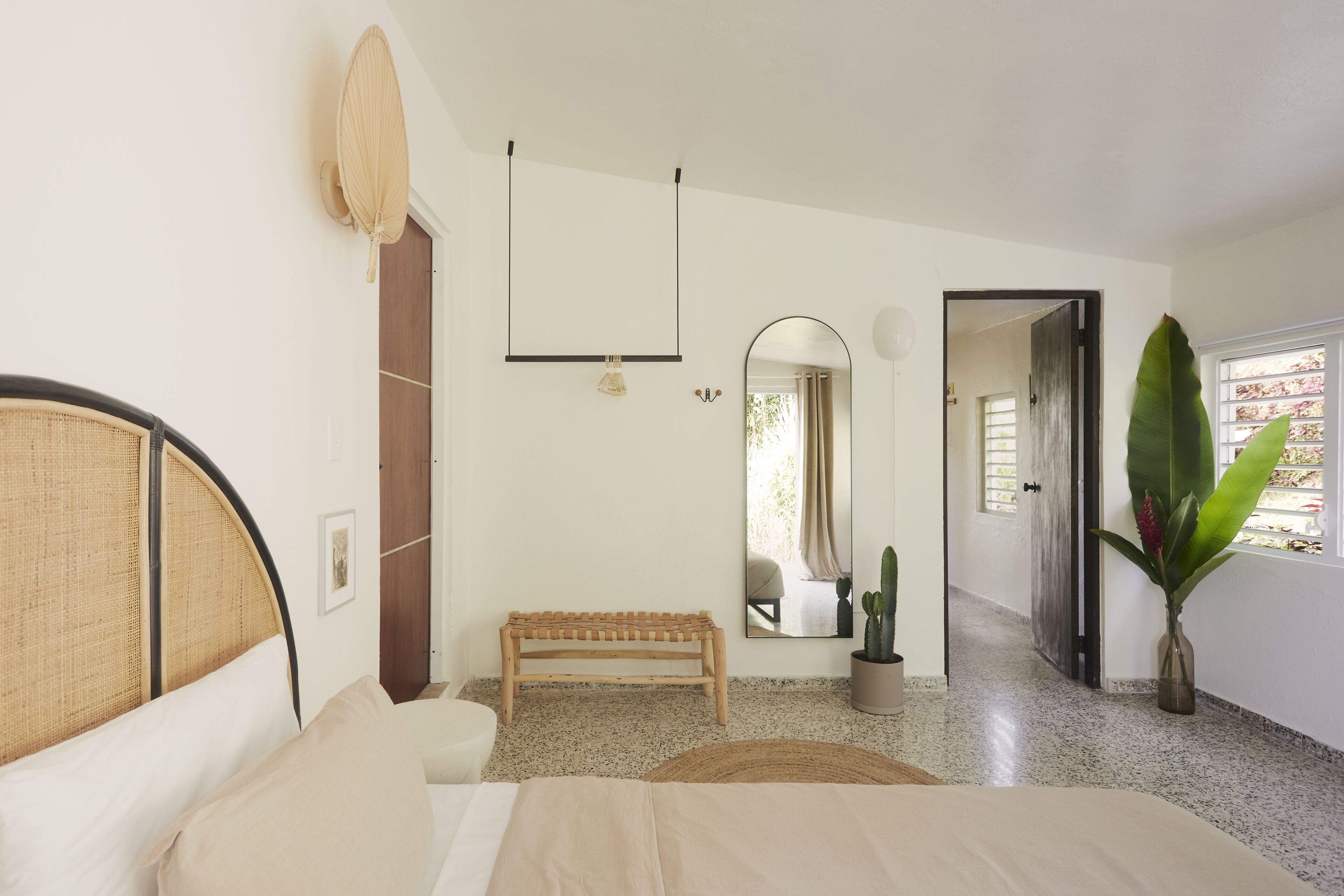 Um quarto com cama, um banco, um espelho, uma planta em vaso e uma porta que dá para outro cômodo.