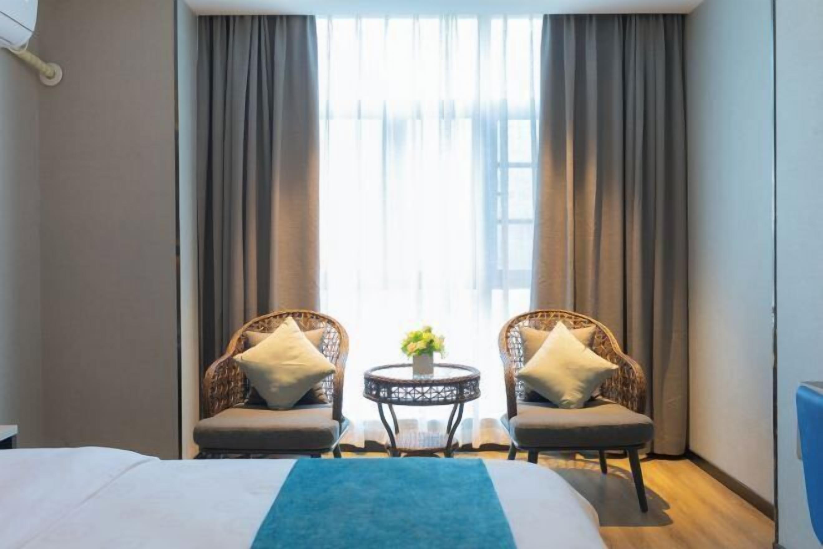 Quarto de hotel com cama, duas cadeiras de vime, uma mesa redonda pequena com um vaso e vista da cidade através de janelas amplas.