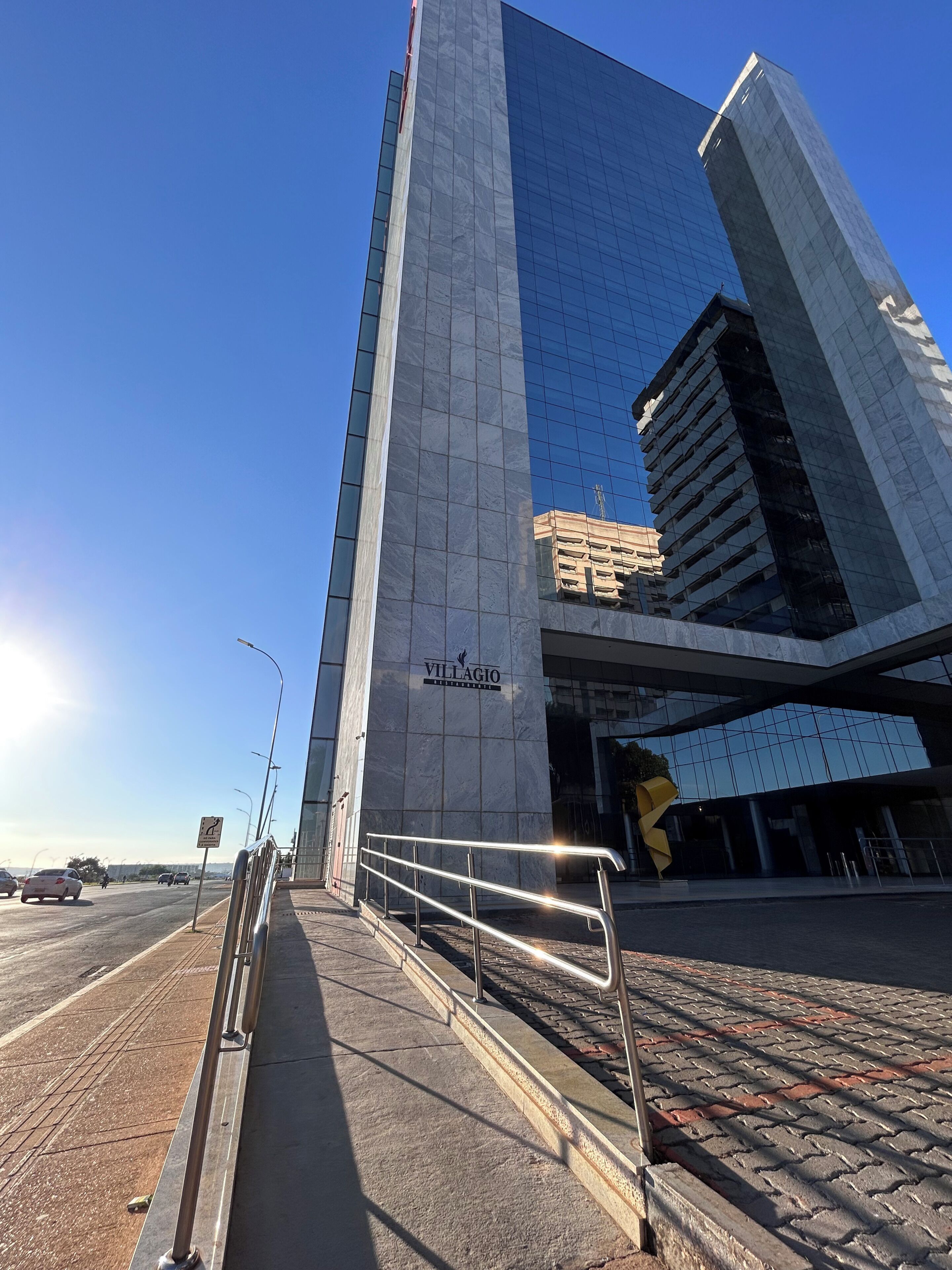 Um edifício moderno com uma placa grande 'VILLAGIO', uma passarela para pedestres e um céu claro.