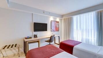 Quarto de hotel com cama, secretária com cadeira, televisão e janela com cortinas.
