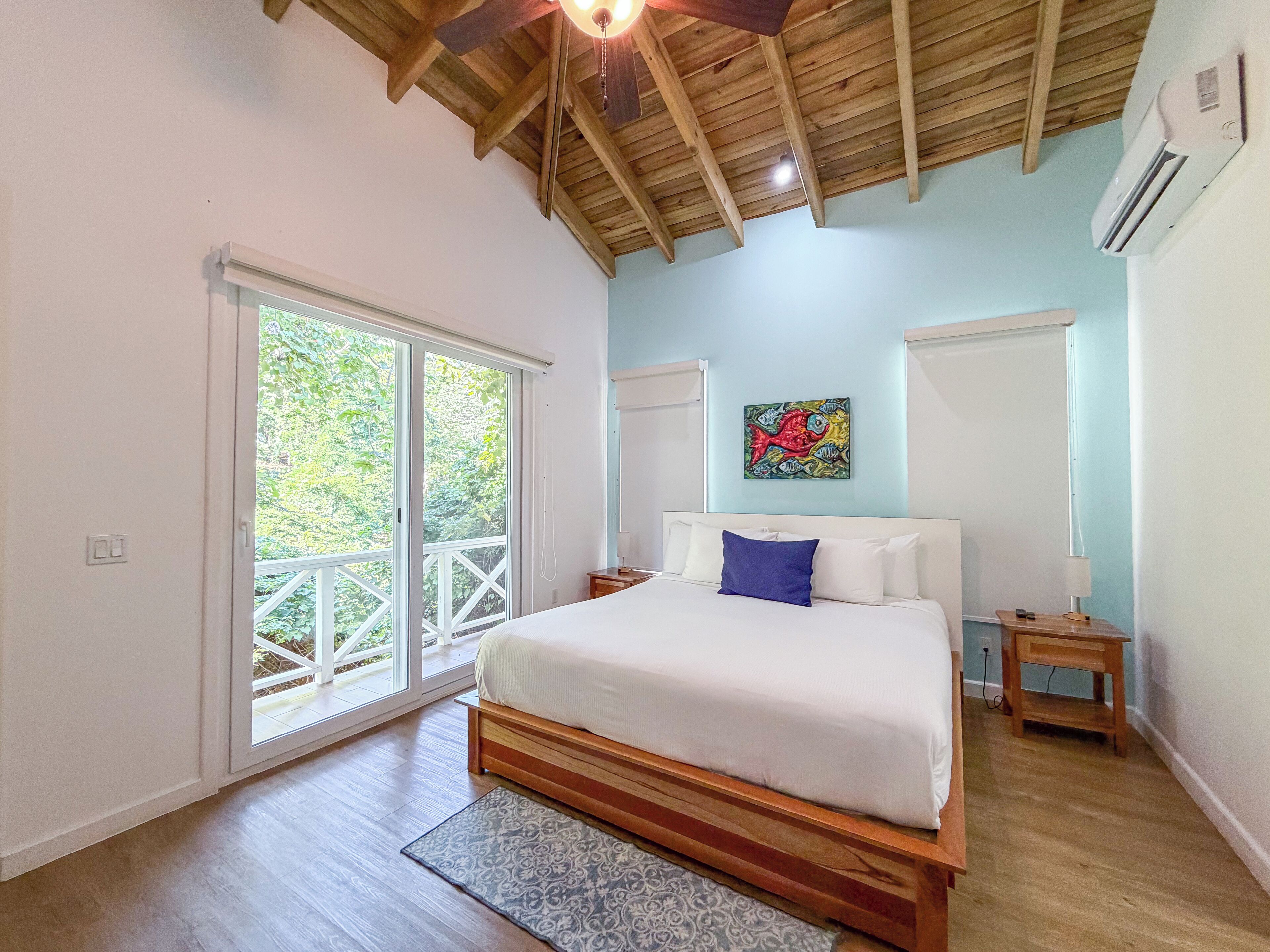 Quarto com teto de madeira, uma cama grande, mesa de cabeceira e janela com vista para o verde.